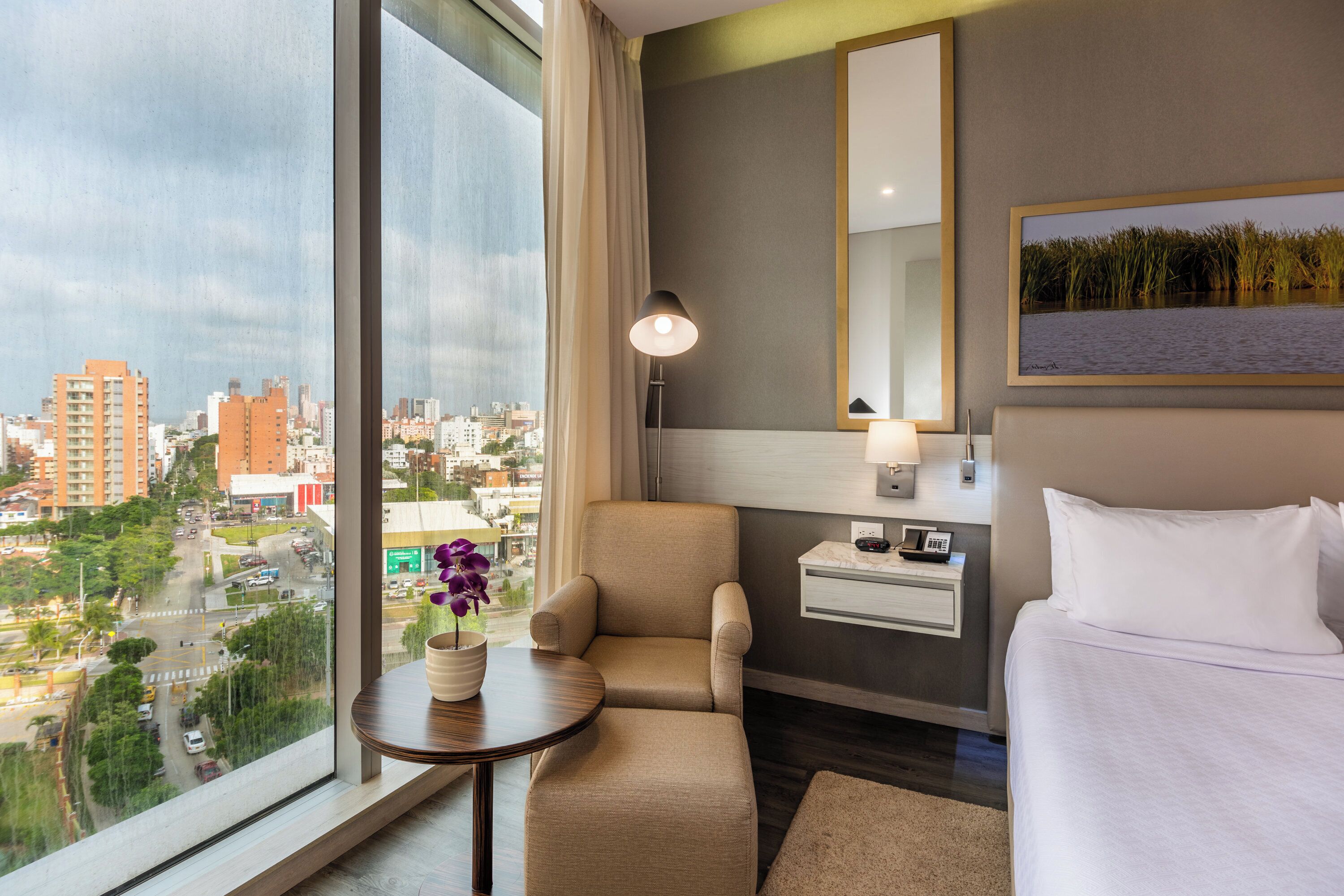 Quarto de hotel com uma janela grande, uma cama, uma cadeira, uma mesinha, uma mesa de cabeceira com abajur e um quadro.