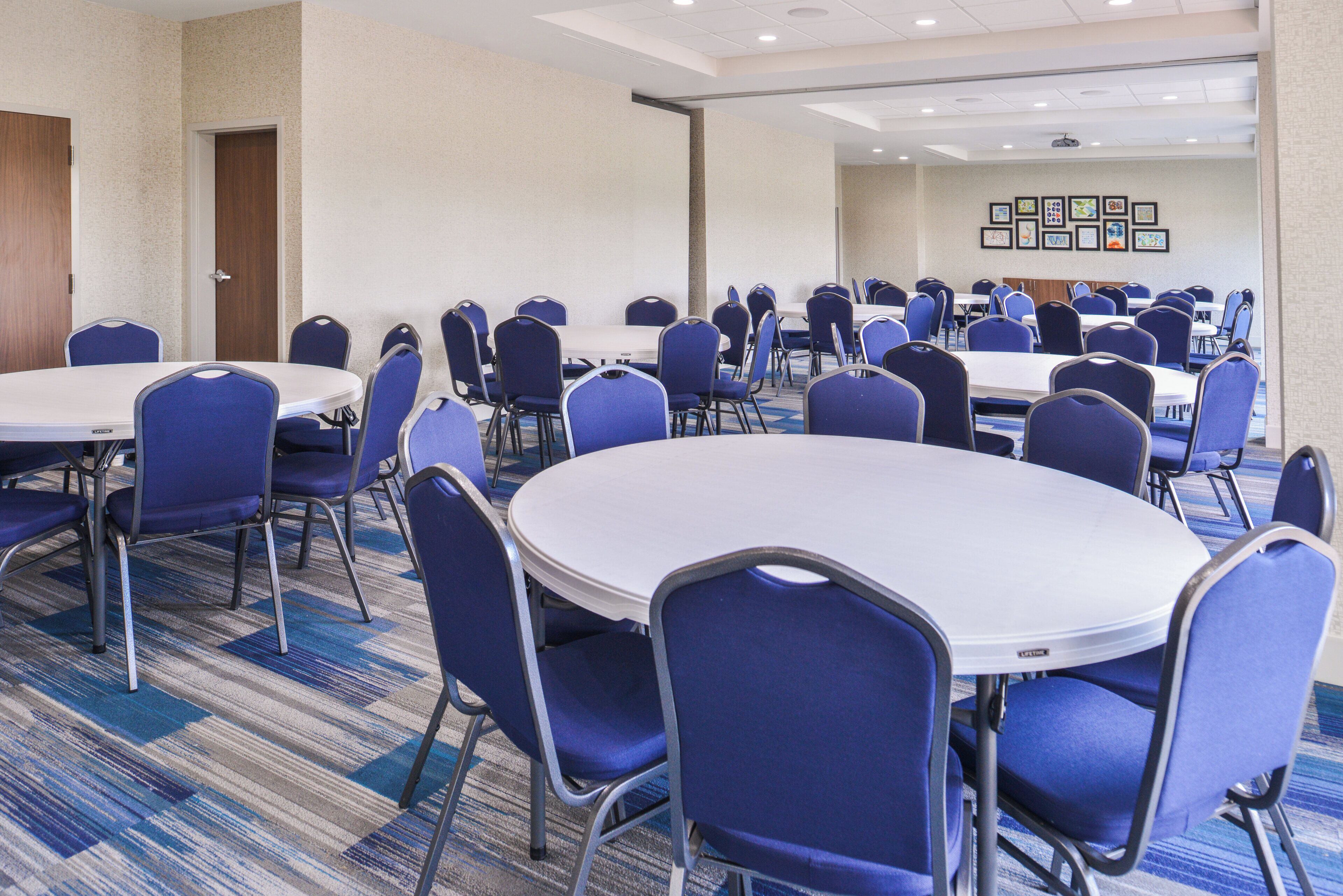 Sala de conferências com mesas redondas e cadeiras azuis, piso carpeteado e quadros nas paredes.