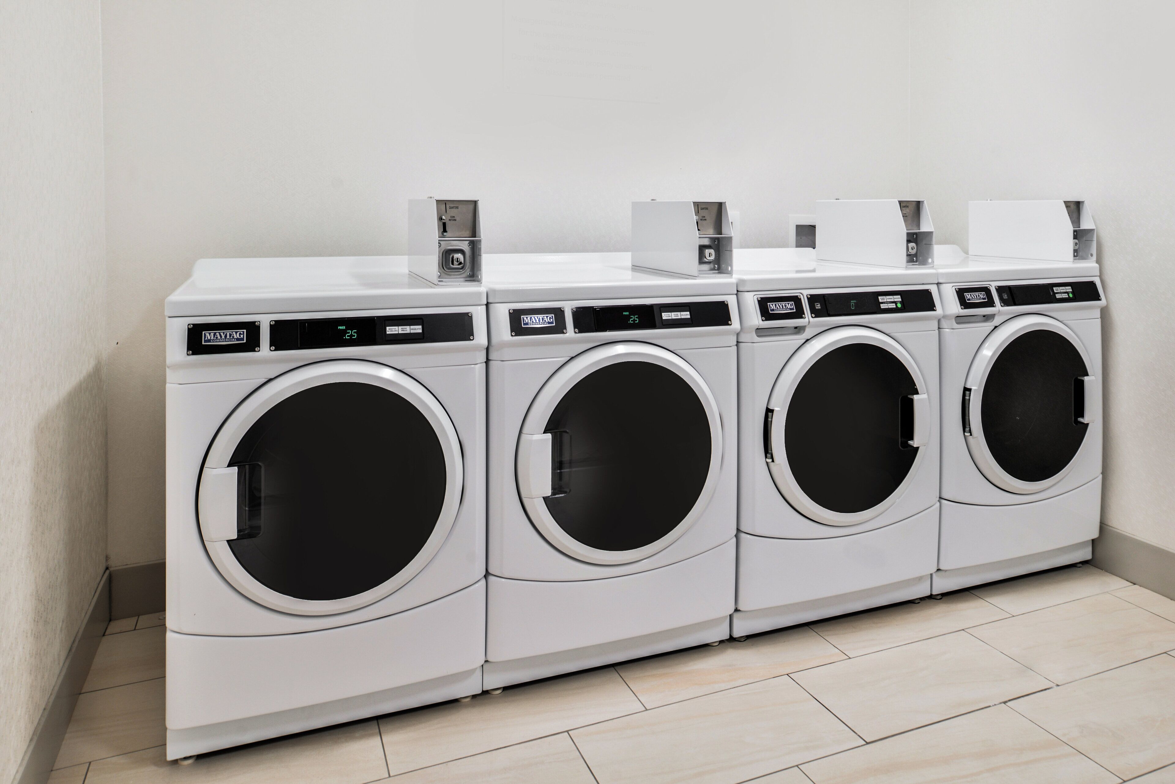 Quatro máquinas de lavar roupa brancas, do tipo frontal, dispostas em linha em um ambiente comercial.