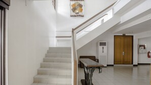 Escada que dá acesso a um segundo andar, uma mesa de madeira e uma porta em um espaço interno limpo e branco.