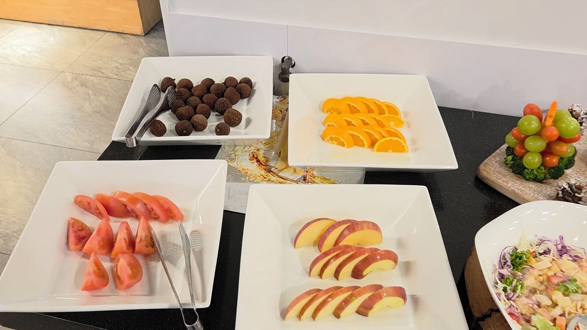 Uma mesa de buffet com diversos alimentos, incluindo fatias de maçã, laranjas e uma salada.