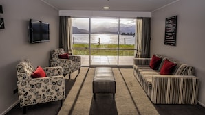Sala moderna com uma grande janela que oferece vista para um lago e montanhas. Há dois sofás e um banco com almofadas vermelhas.