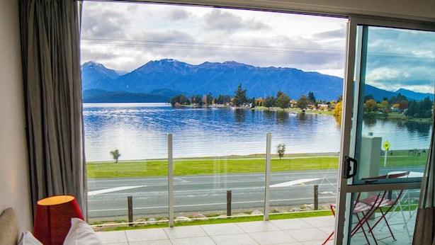 Quarto com cama grande, vista para um lago e montanhas, e varanda com uma cadeira.