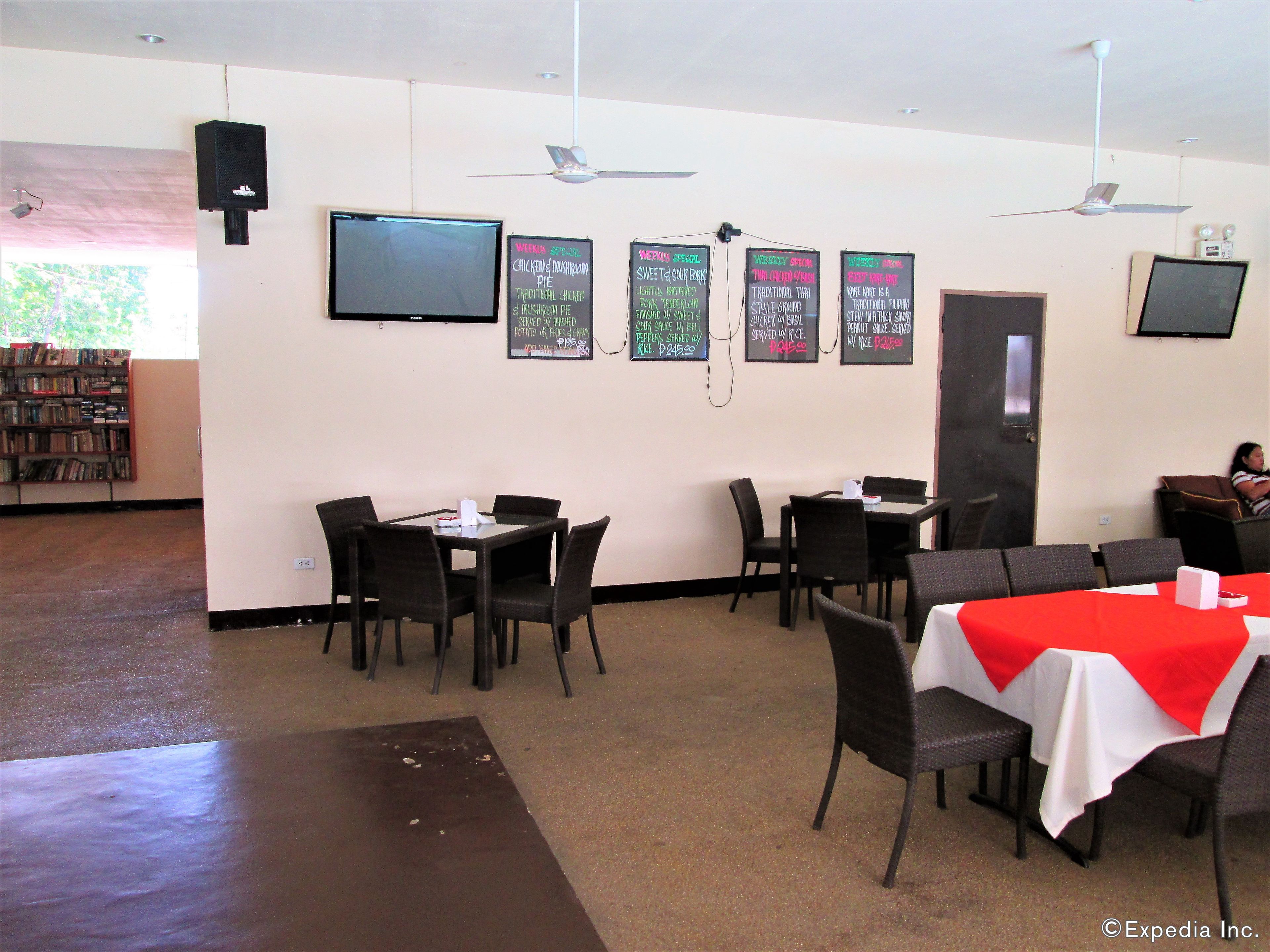Restaurante com várias mesas e cadeiras, televisão fixada na parede e cardápios em lousa exibindo diversos pratos.