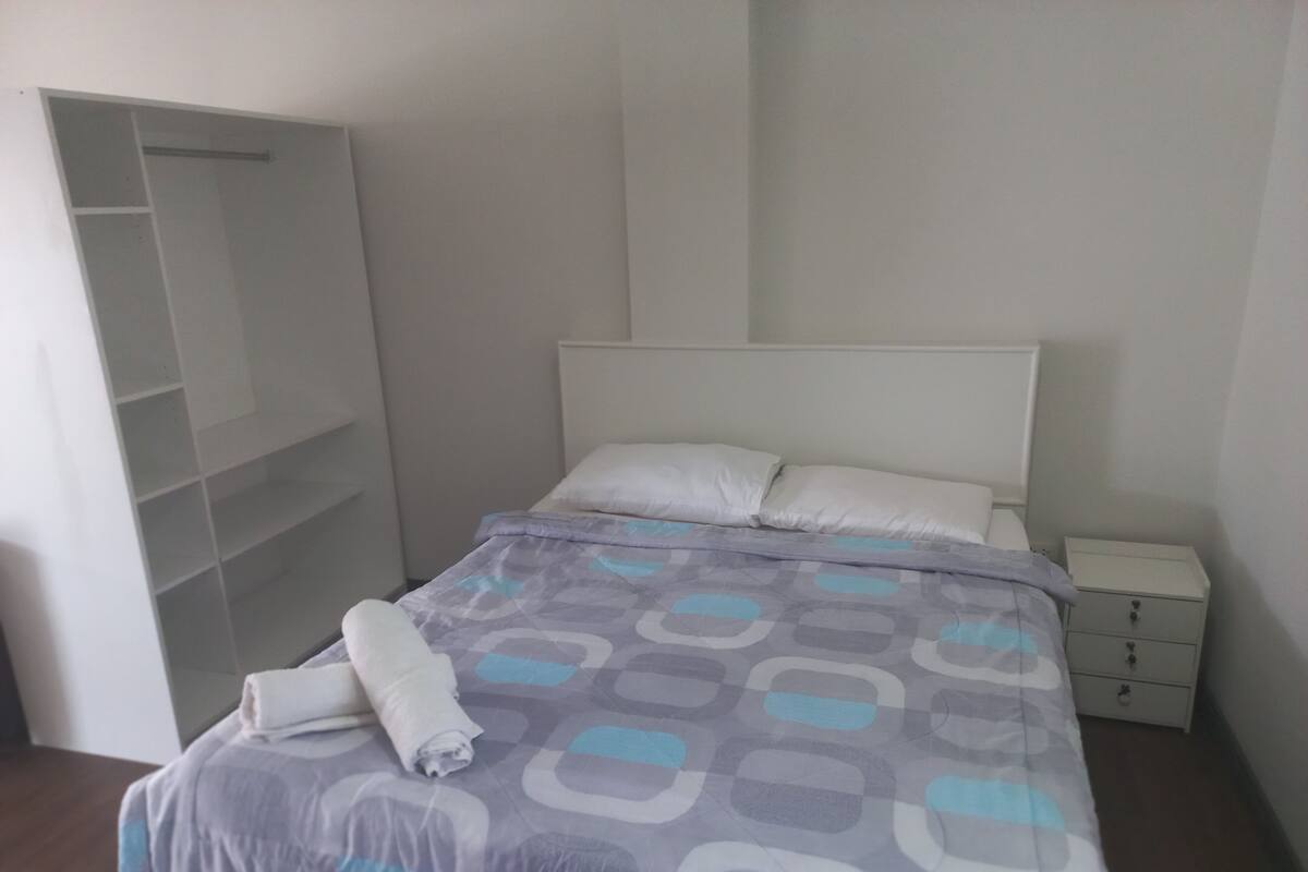 Cama bem arrumada com colcha estampada, cabeceira branca e mesa de cabeceira com gavetas.