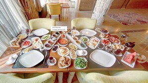 Mesa de jantar posta com diversos pratos e temperos, rodeada por cadeiras em um ambiente bem iluminado com piso de azulejos e um pilar de pedra.