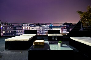 Terraço no telhado com assentos e uma mesa, com vista para a paisagem urbana ao entardecer.