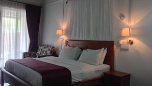 Quarto de hotel com uma cama grande, duas luminárias de cabeceira, uma mesinha de cabeceira e uma janela com cortinas.