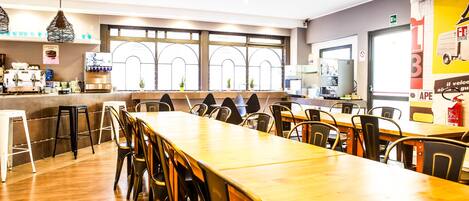 Um restaurante com uma longa mesa de madeira e cadeiras, uma área de bar com máquina de café e grandes janelas com arcos.