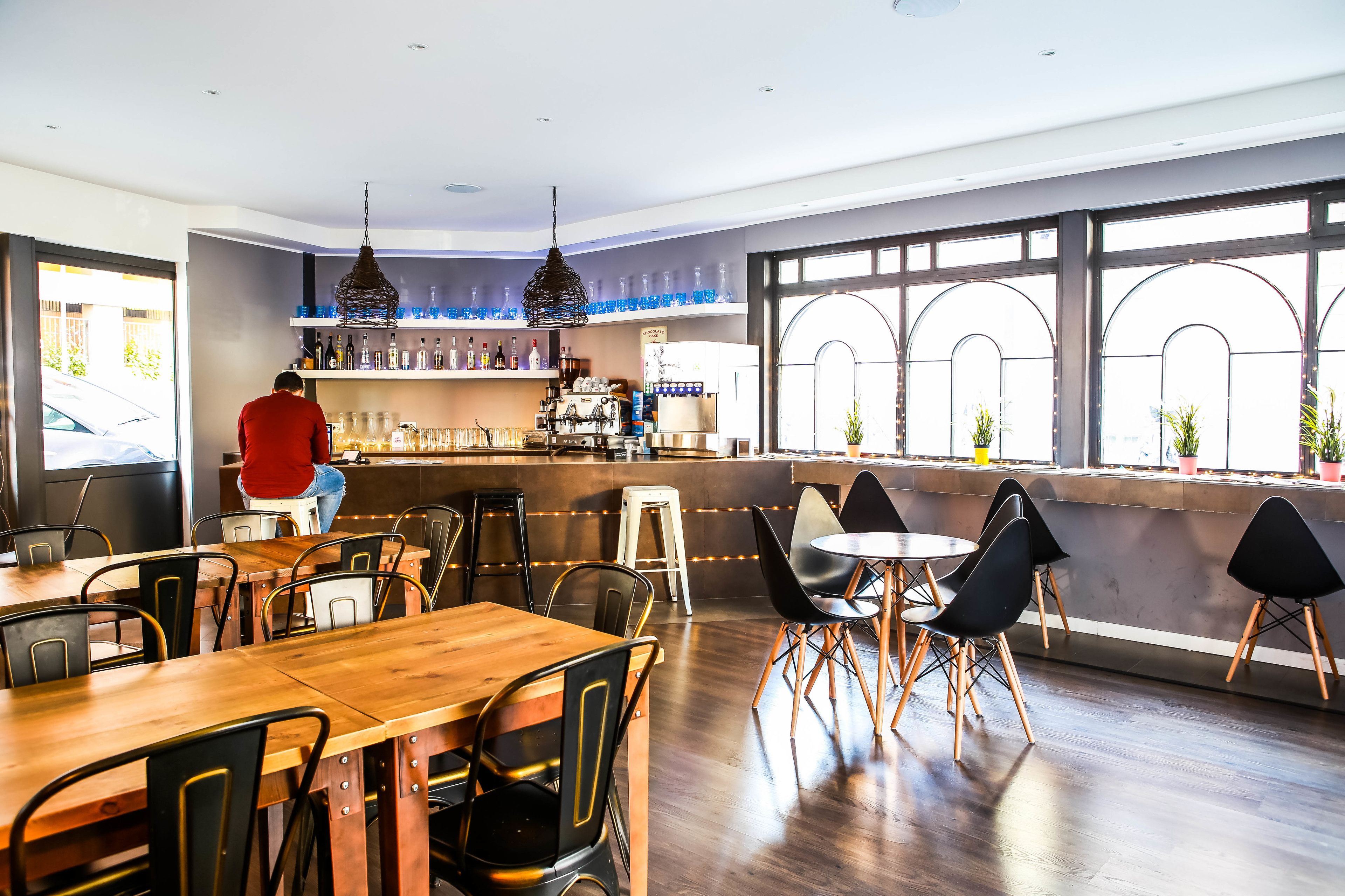 Interior moderno de restaurante com mesas e cadeiras de madeira, área de bar com prateleiras cheias de garrafas, e janelas grandes com arcos.