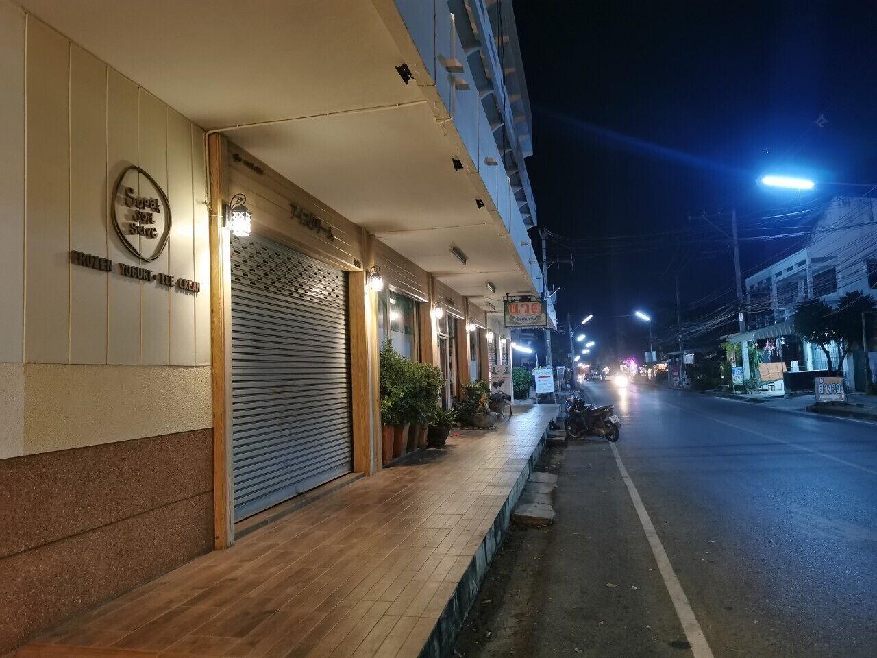 Vista da rua à noite, com uma loja fechada à esquerda e uma moto estacionada na calçada.