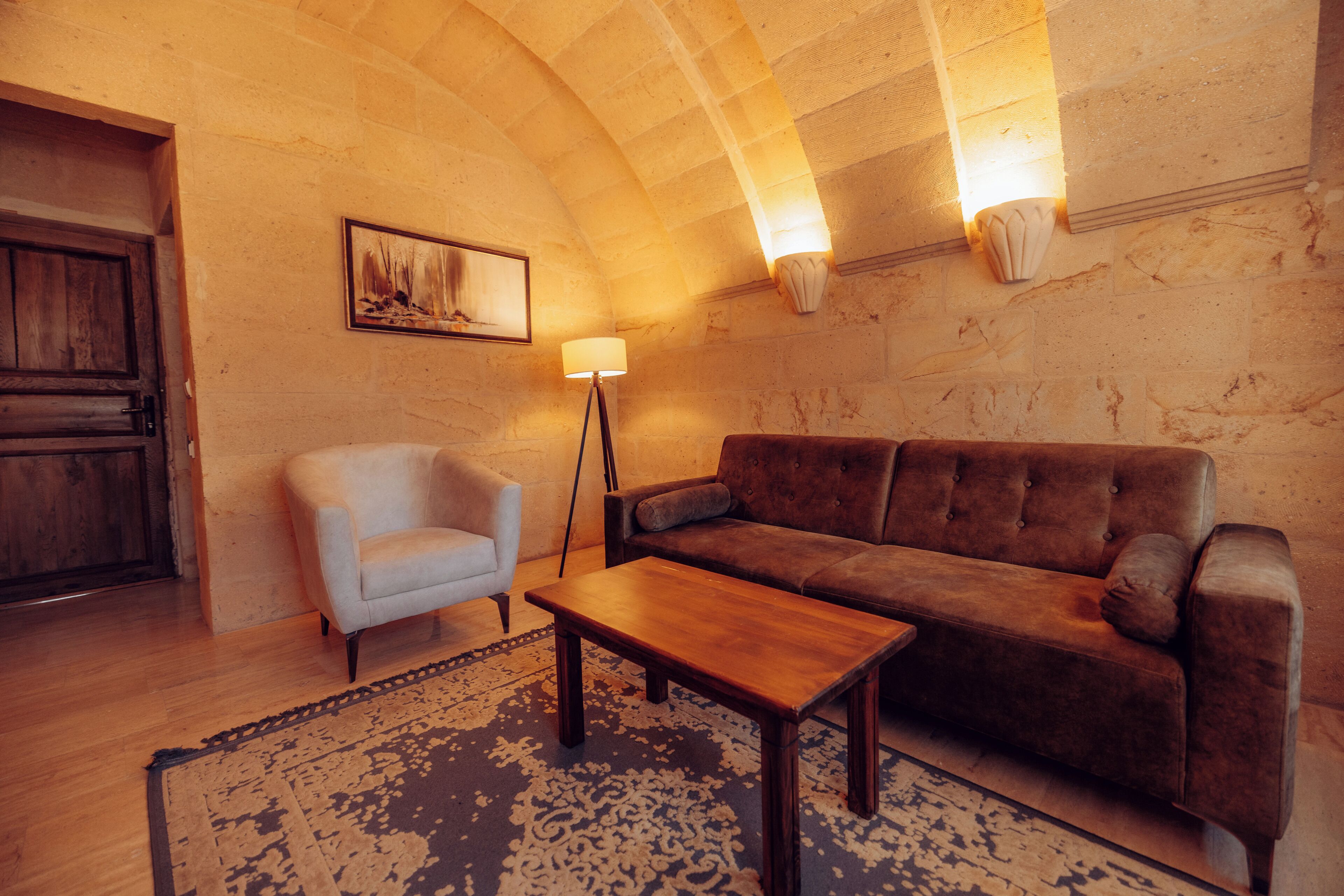Um quarto acolhedor com piso de madeira, um sofá marrom, uma poltrona branca, uma mesa de centro de madeira, um tapete estampado, um quadro na parede e um abajur.