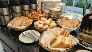Disposição de buffet com diversos pães e doces, máquinas de café e utensílios para refeições.