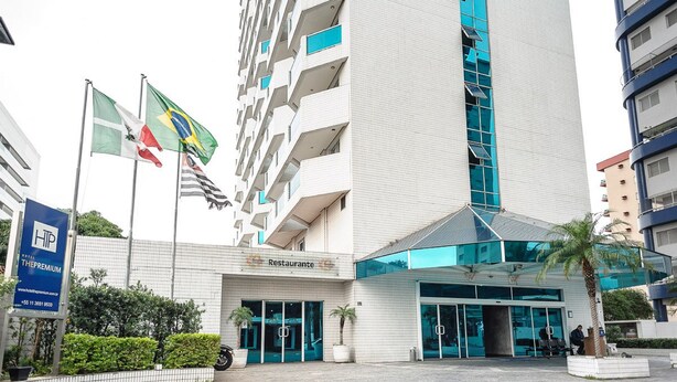 Entrada de hotel com fachada de vidro, uma placa com 'The Premium Hotel' e várias bandeiras, incluindo a bandeira do Brasil.