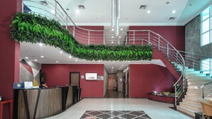 Um saguão de hotel com balcão de recepção, escada e parede verde com plantas.