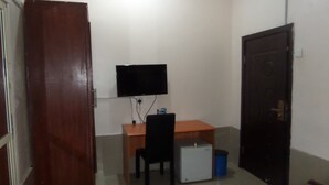 Um quarto pequeno com uma escrivaninha de madeira, uma cadeira, uma televisão na parede, um armário pequeno e uma porta.