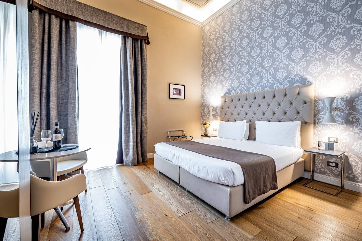 Quarto de hotel com cama, mesa de cabeceira, luminária e escrivaninha com cadeiras.