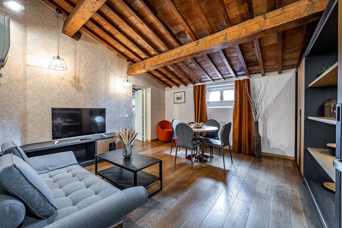 Uma sala de estar moderna com sofá, mesa de centro e televisão. Há uma área de jantar com mesa e cadeiras. O aposento possui vigas de madeira no teto e uma grande janela com cortinas.