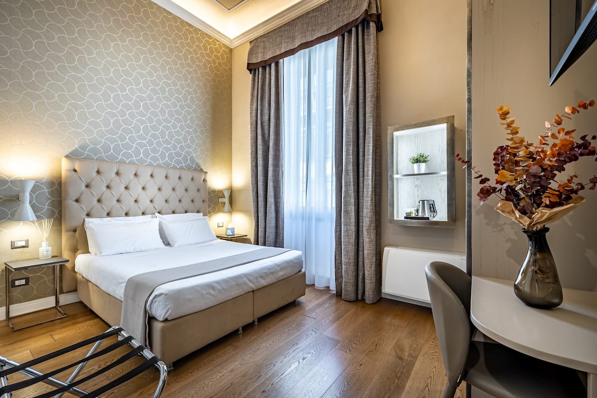 Quarto de hotel moderno com uma cama grande, mesa de cabeceira com abajur, prateleira pequena com uma planta e um vaso com flores.