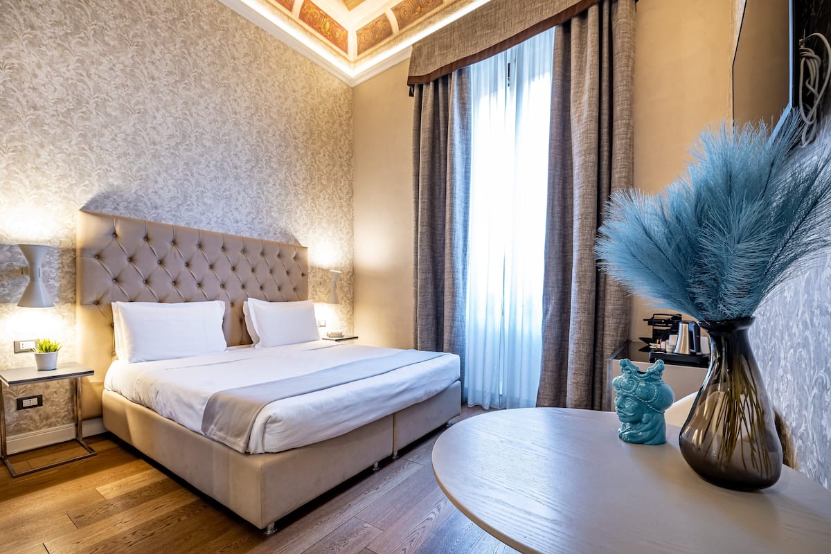 Quarto de hotel com cama de cabeceira estofada, mesa de cabeceira com abajur, vaso com penas azuis e mesa redonda.