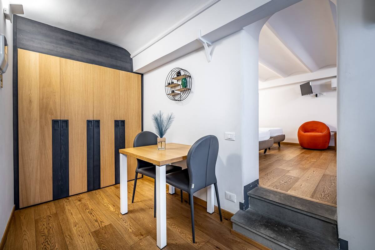 Uma área de refeições com uma mesa e cadeiras de madeira, uma estante fixada na parede com objetos decorativos, e uma escada que leva a outro cômodo.
