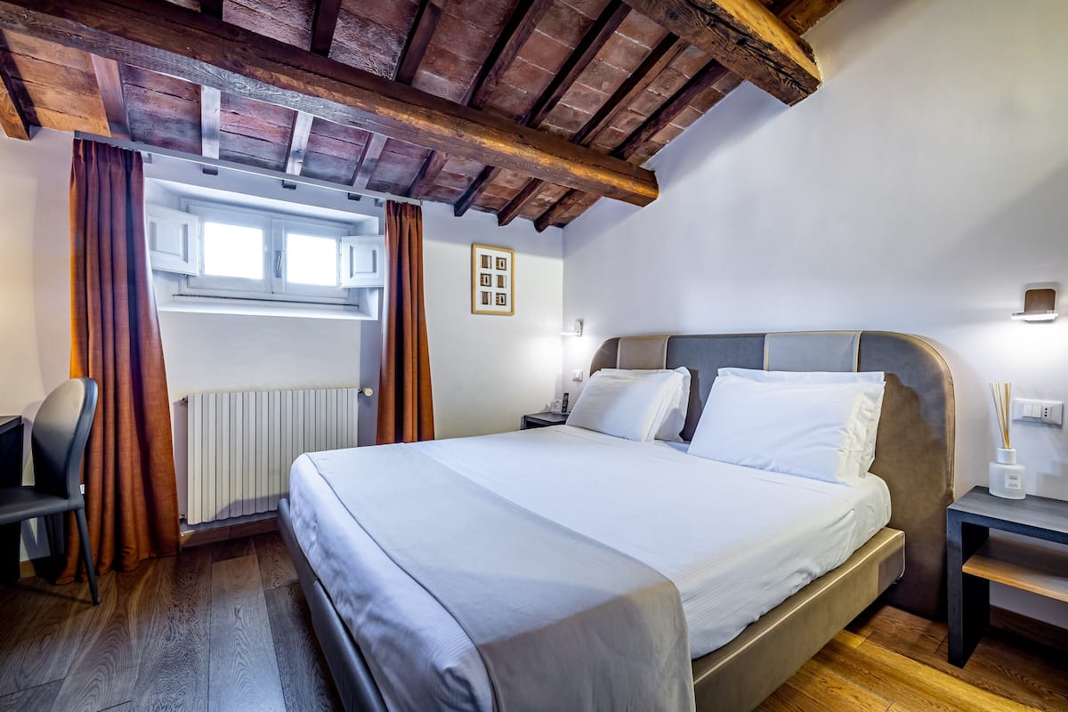 Quarto de hotel com uma cama grande, vigas de madeira no teto, um radiador, uma escrivaninha e uma janela com cortinas.