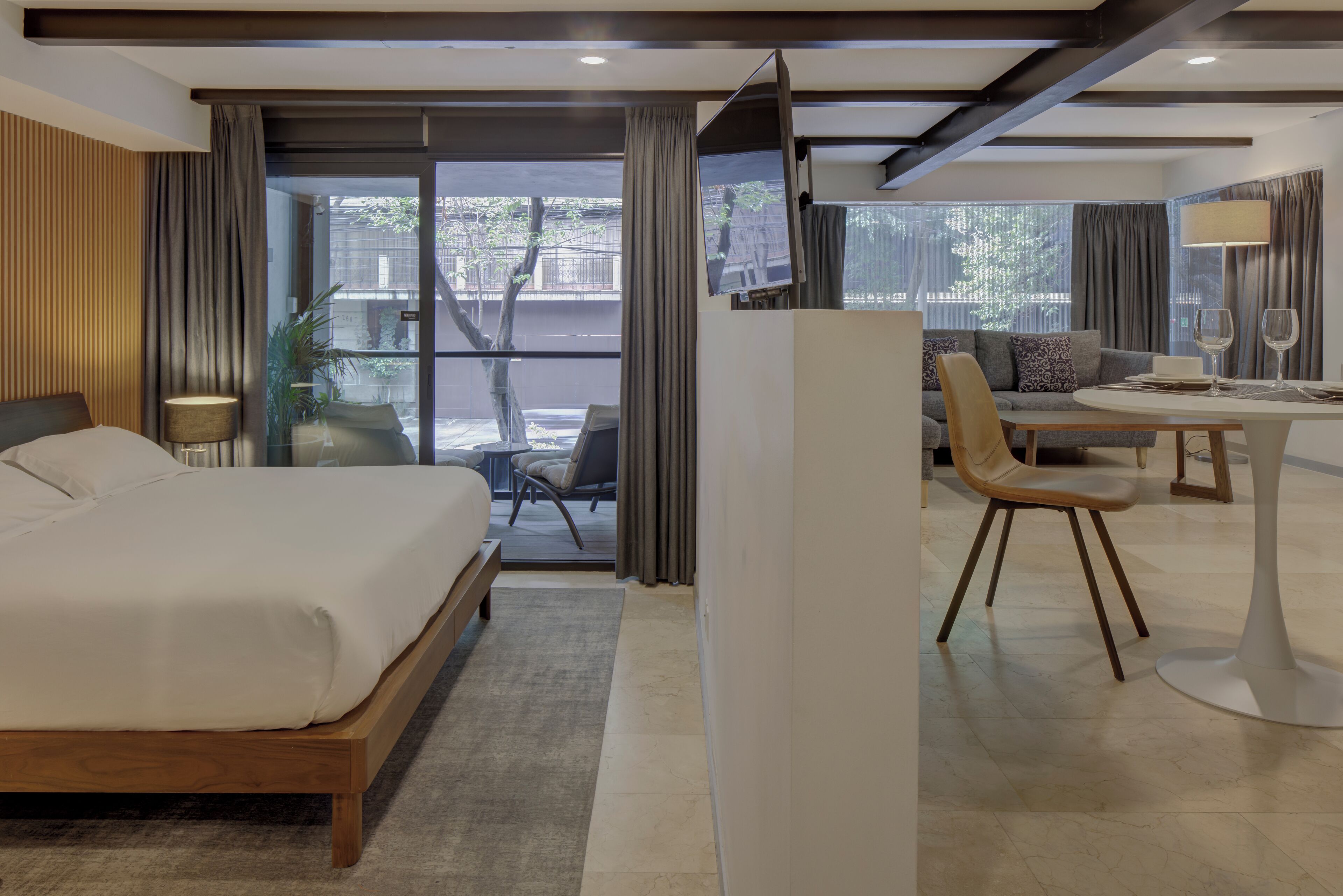 Quarto de hotel moderno, com uma cama grande, área de jantar com mesa e cadeiras, e vista através de uma porta de vidro.