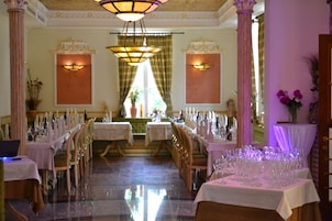 Uma sala de jantar formal com várias mesas postas para refeição, elegantes lustres e arandelas decorativas.