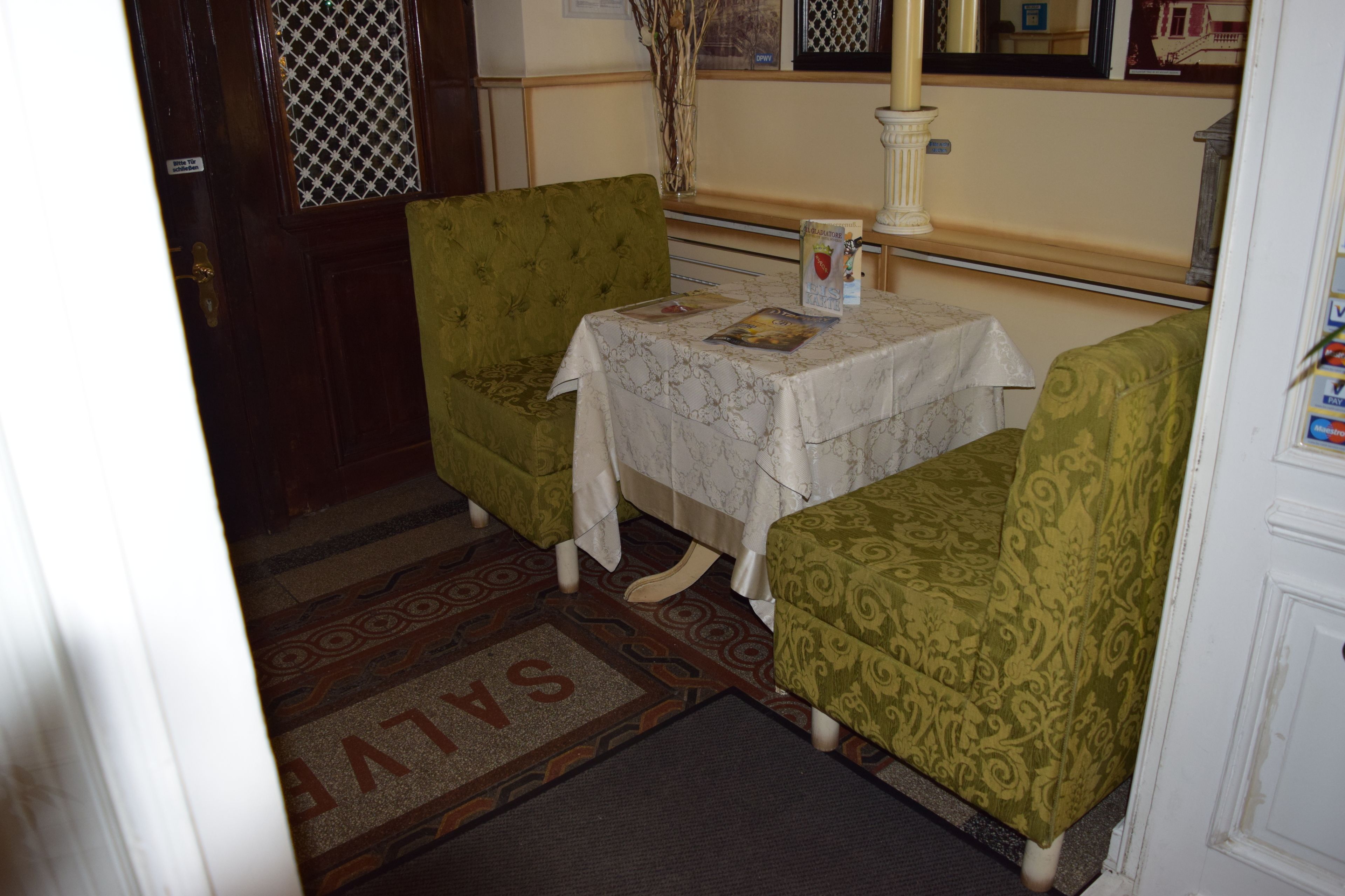 Um quarto com duas poltronas verdes estofadas, uma mesa pequena com toalha branca e um tapete estampado com a palavra "SALUTE".