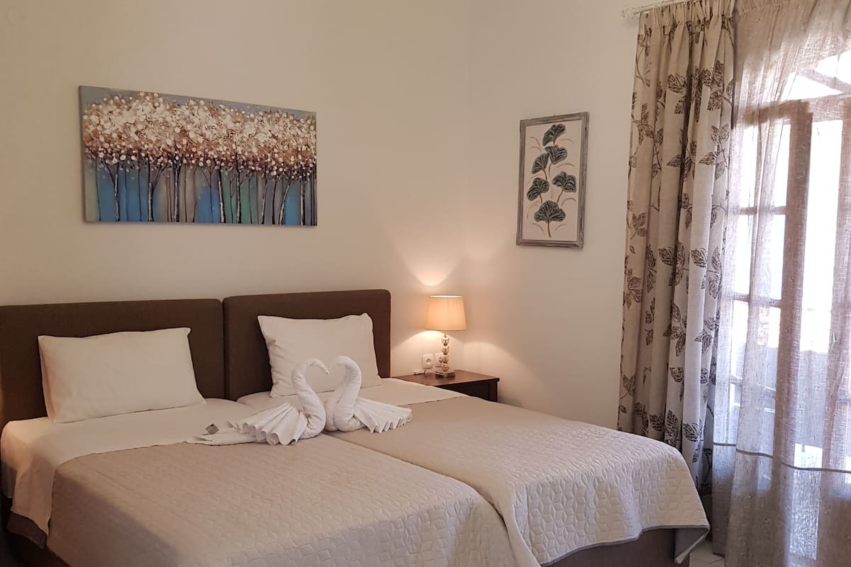Uma cama de casal com colcha acolchoada, uma mesinha de cabeceira com abajur e duas pinturas penduradas na parede.
