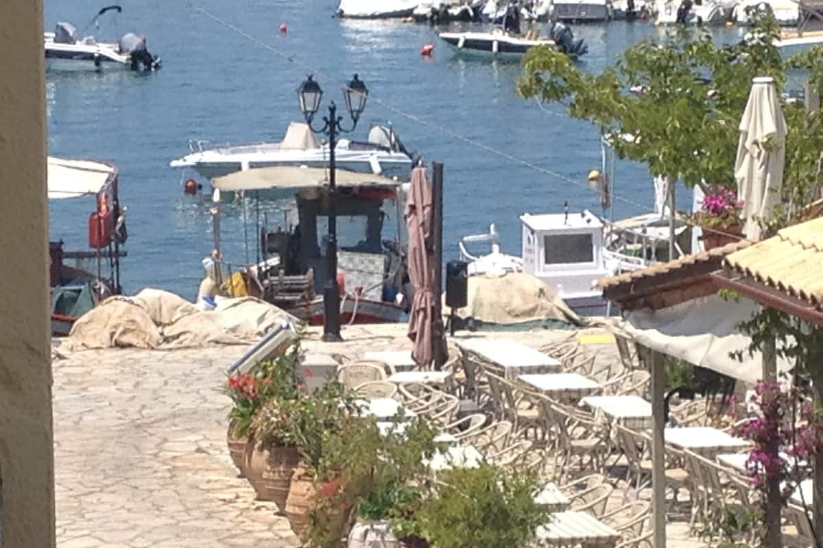 Uma área costeira com barcos atracados, calçada de pedra, mesas ao ar livre e um gazebo.
