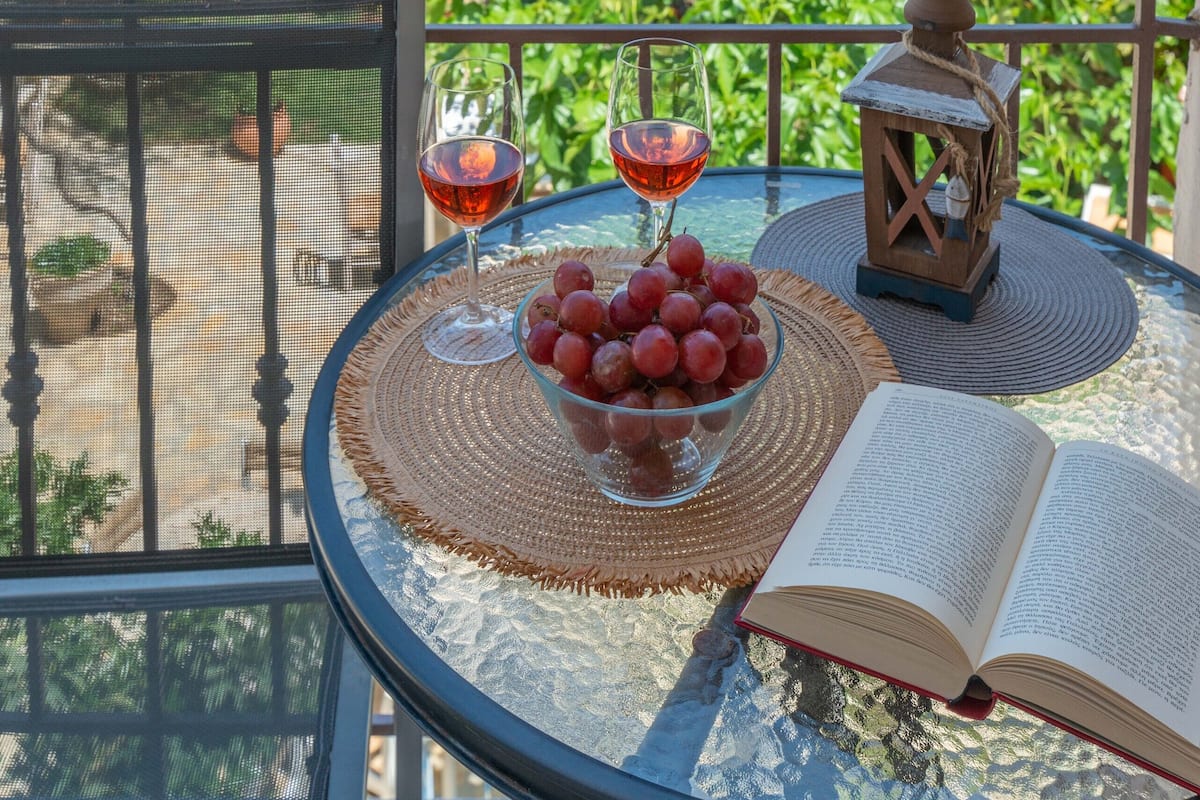 Varanda com mesa posta para dois, com uma tigela de uvas, duas taças de vinho, um candeeiro e um livro aberto.