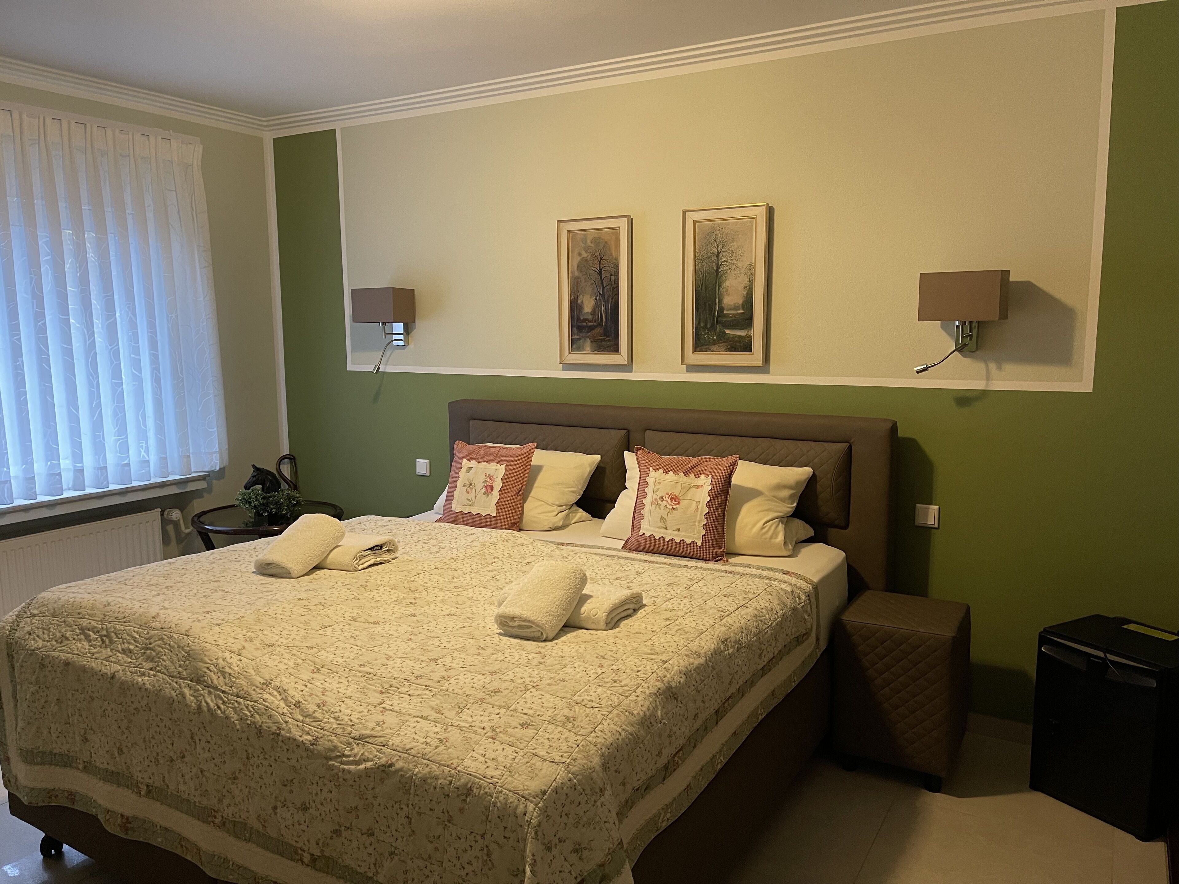 Uma cama bem arrumada com colcha florida, duas pinturas emolduradas de paisagens na parede e um abajur.