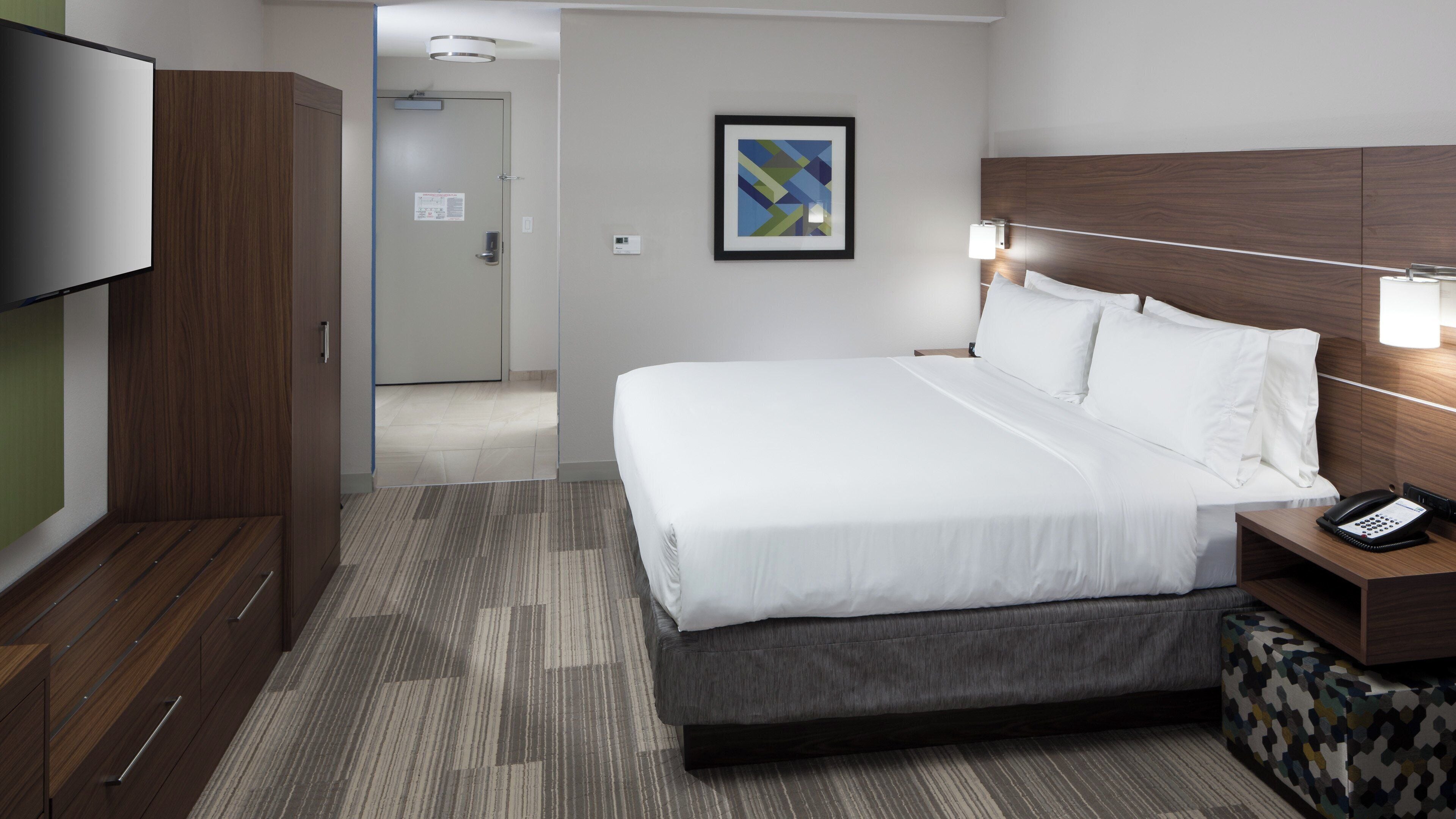 Quarto de hotel com cama, mesa de cabeceira, abajur e televisão à esquerda.