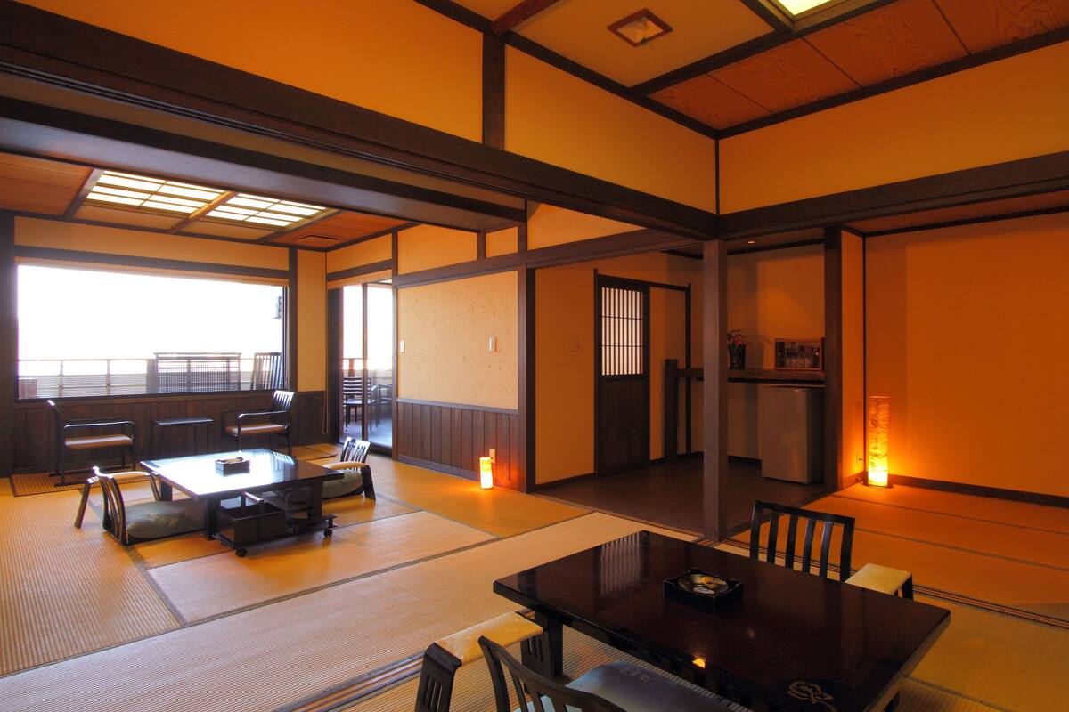 Um quarto japonês tradicional com tatames, uma mesa baixa e cadeiras. Há uma porta deslizante e uma pequena cozinha.