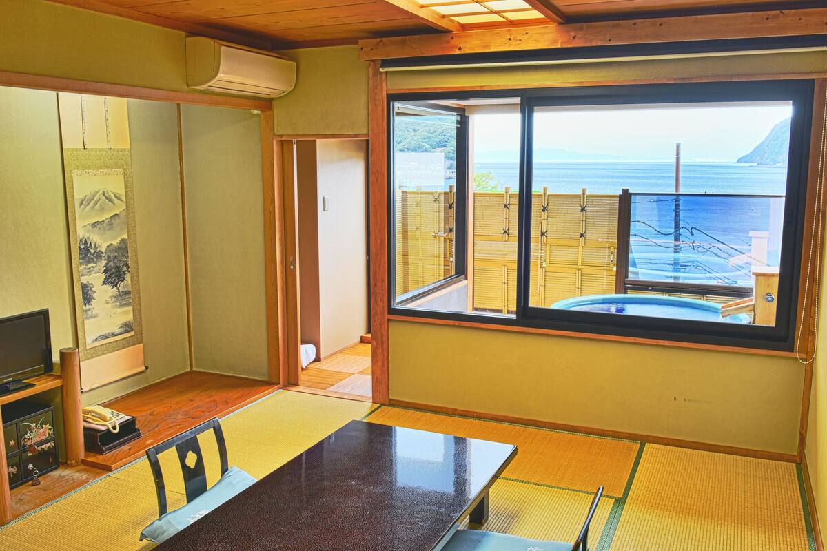 Um quarto japonês tradicional com piso de tatame, uma mesa de tatame, uma mesa de madeira baixa, uma televisão e vista para o mar através de uma grande janela.