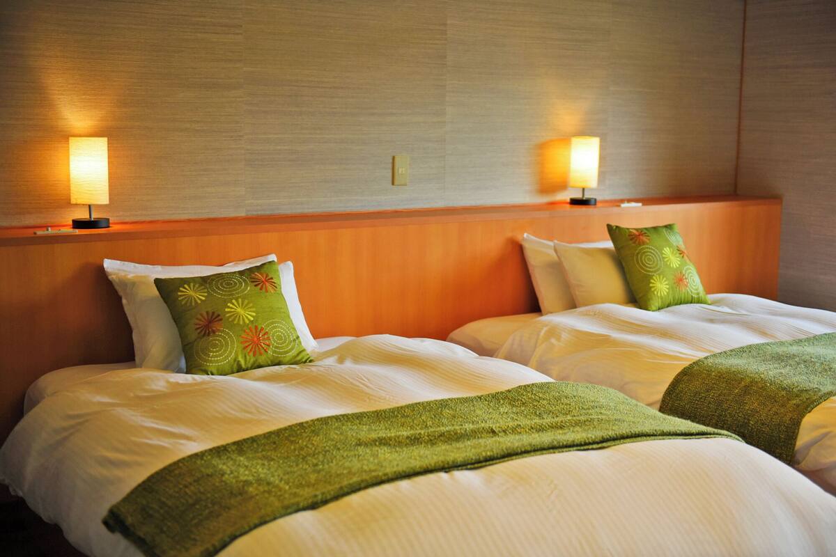 Quarto de hotel com duas camas, cada uma com roupa de cama branca e mantas verdes. Duas luminárias fixadas na parede fornecem iluminação.