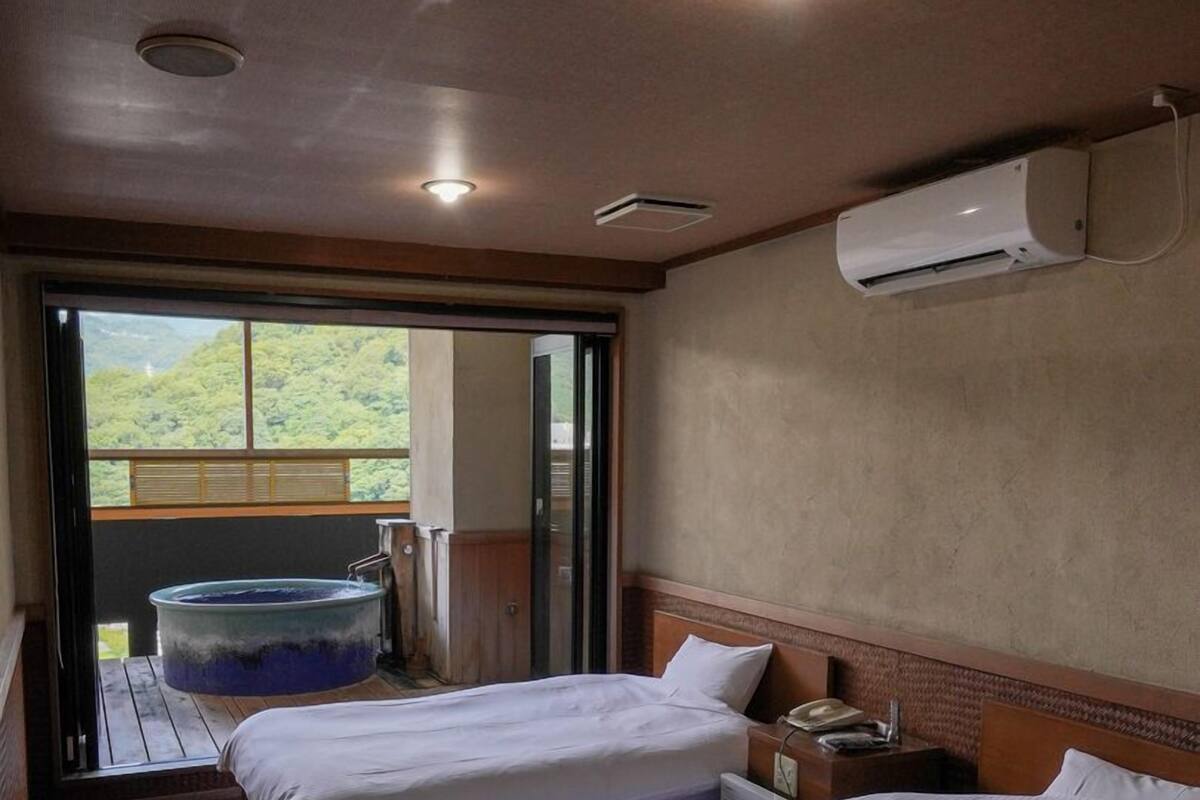 Quarto com duas camas, piso de madeira, aparelho de ar condicionado e vista para o verde lá fora.