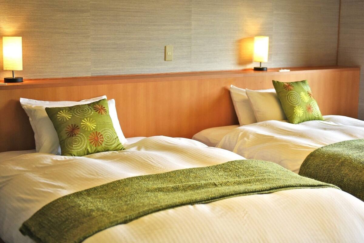Quarto de hotel com duas camas, cada uma com travesseiro verde, cabeceira de madeira e duas luminárias fixadas na parede.