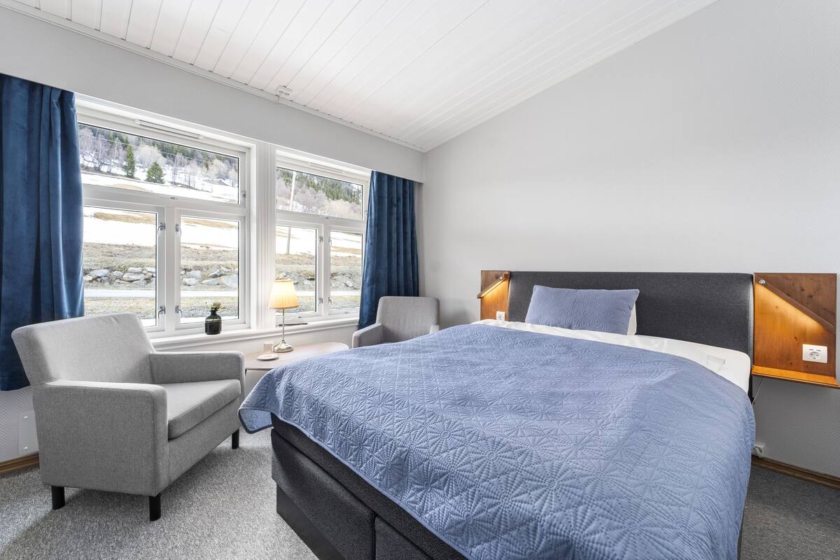 Quarto de hotel com uma cama grande, uma cadeira, uma escrivaninha e uma janela com cortinas.
