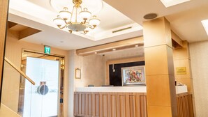 Uma área de recepção de hotel com design moderno, apresentando um balcão de atendimento, um candelabro e uma porta de vidro com sinalização de saída.
