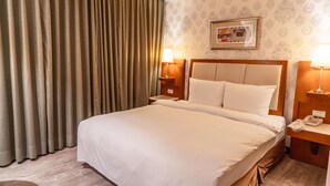 Quarto de hotel com uma cama grande, duas mesas de cabeceira com luminárias, um quadro na parede e uma cortina na janela.