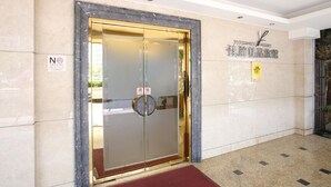 Entrada de um hotel com parede de mármore, porta de vidro emoldurada a dourado e tapete vermelho que leva à entrada.