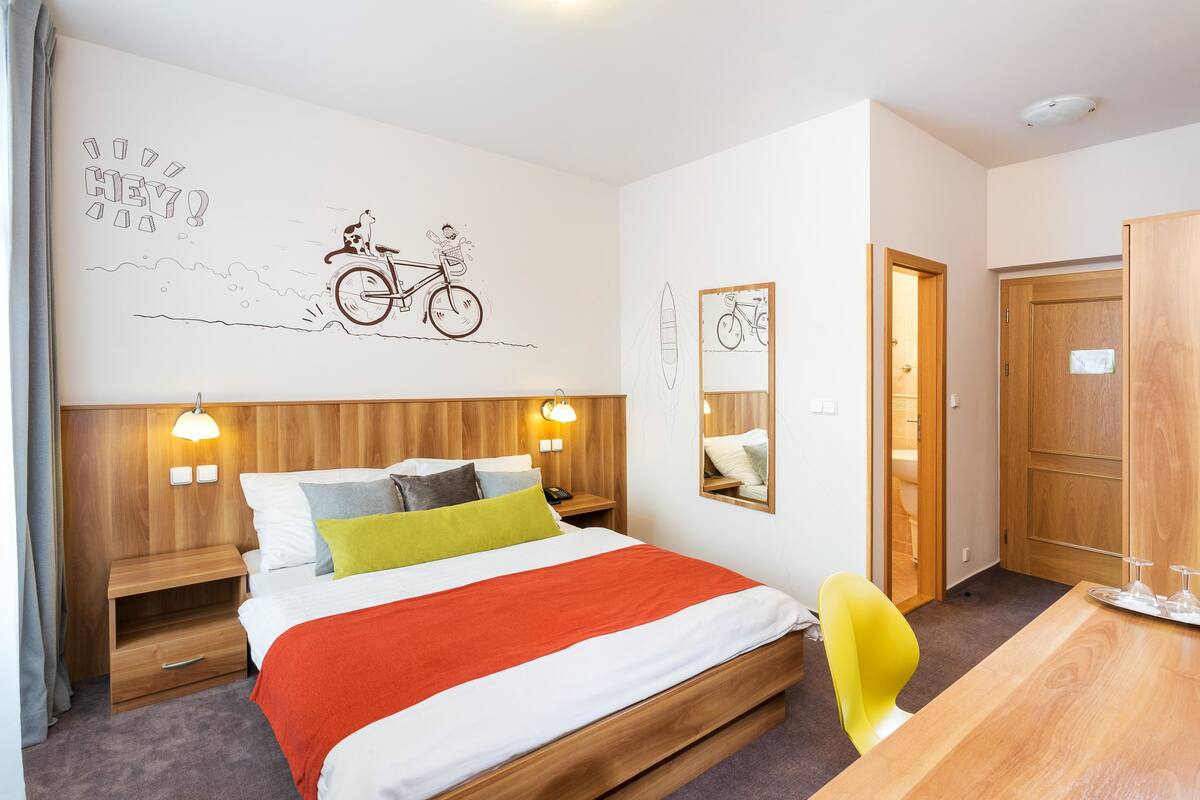 Quarto de hotel com cabeceira de madeira, cama com roupa de cama branca e vermelha, espelho, mesa de cabeceira de madeira, escrivaninha com cadeira e parede com desenho de bicicleta e texto.