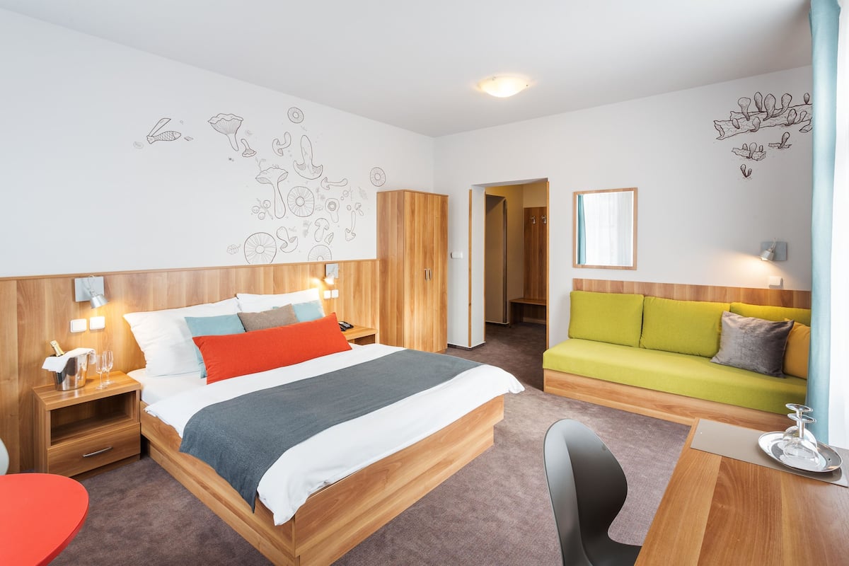 Quarto de hotel moderno com cama de madeira, sofá verde, mesa de jantar e pequena cozinha.