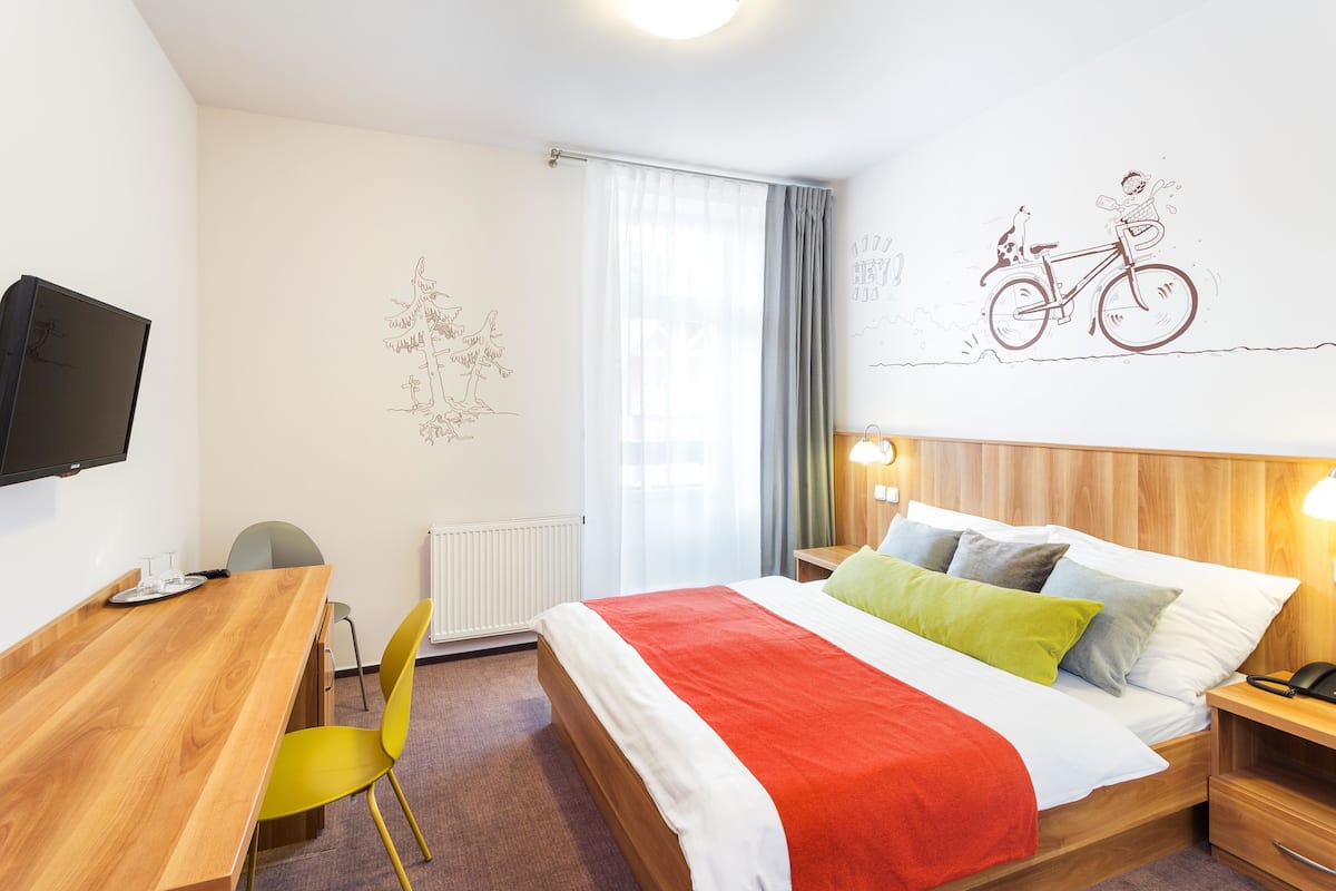 Quarto de hotel com cama de madeira, escrivaninha, cadeira, televisão e janela com cortinas.