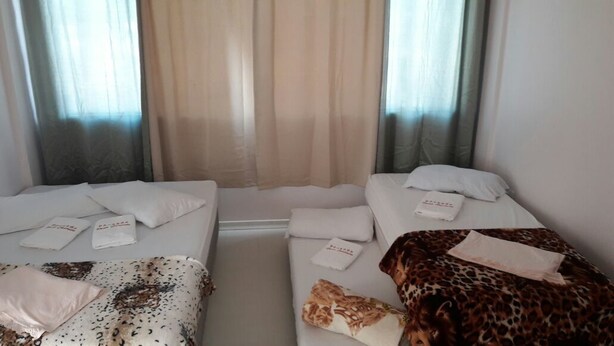 Quarto de hotel com duas camas, cada uma com cabeceira e almofadas brancas. Há cortinas nas janelas e uma pequena mesa com uma luminária entre as camas.