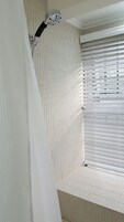 Chuveiro com chuveiro manual, janela com persianas e parede revestida com azulejos.