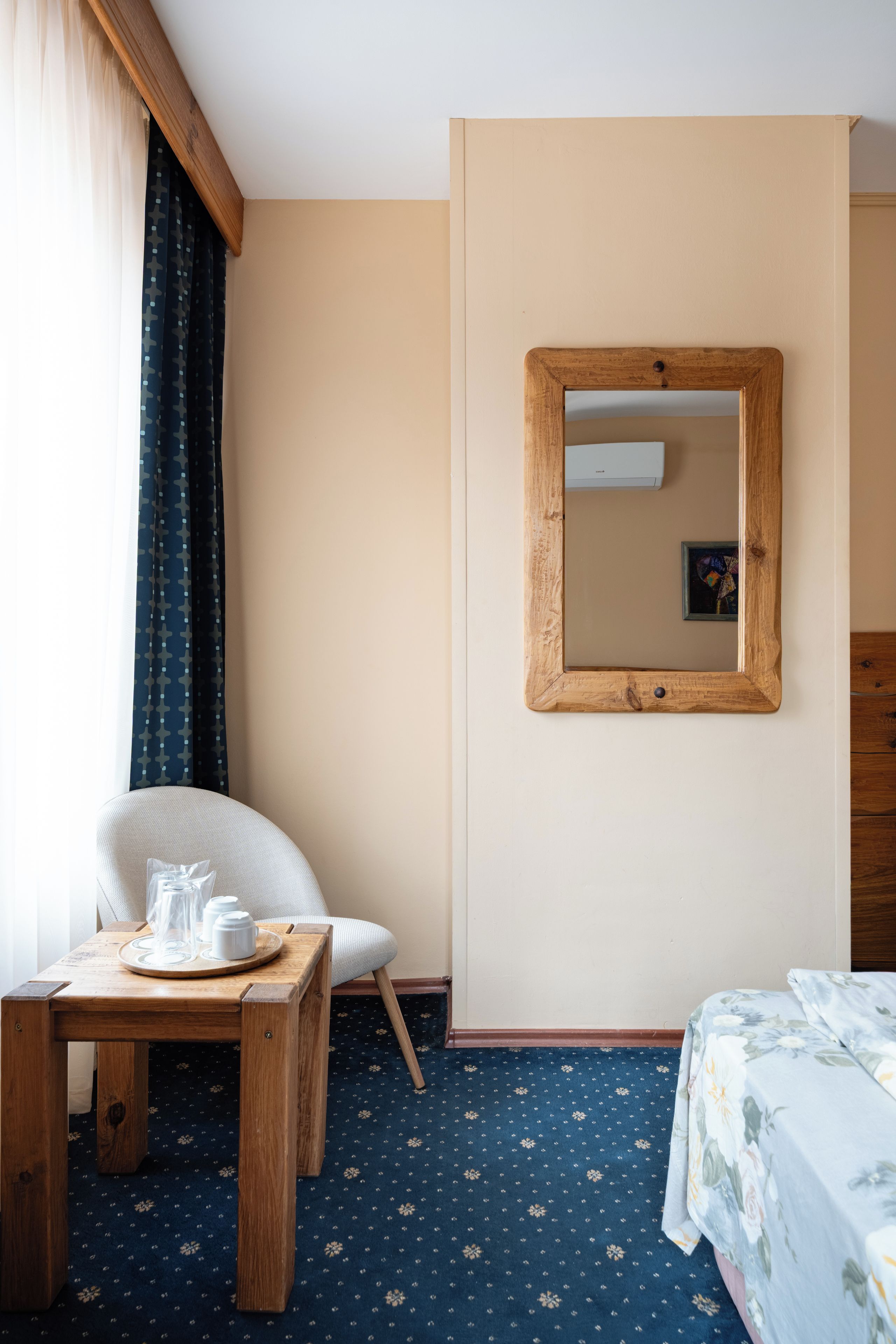 Quarto de hotel com mesa de madeira, uma cadeira, cama com roupa de cama floral, espelho na parede e tapete estampado.