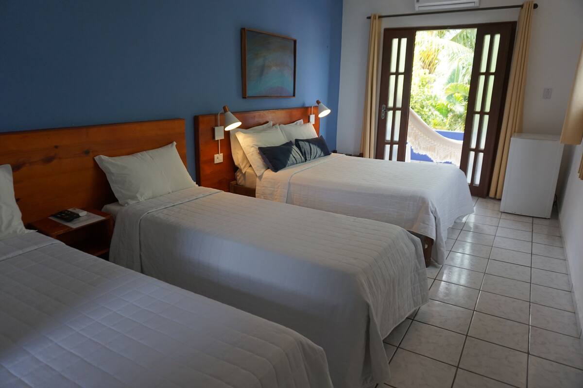 Quarto de hotel com duas camas, cabeceira de madeira, um quadro na parede e vista para o verde através de uma porta.